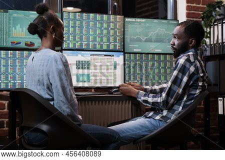 Sales Consultants Sitting At Multi Monitor Workstation While Discussing ...